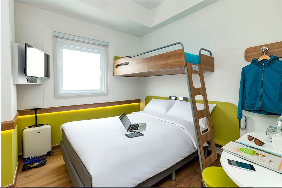 Quarto de hotel com uma cama beliche, uma escrivaninha com cadeira, uma mala, um laptop e uma janela.