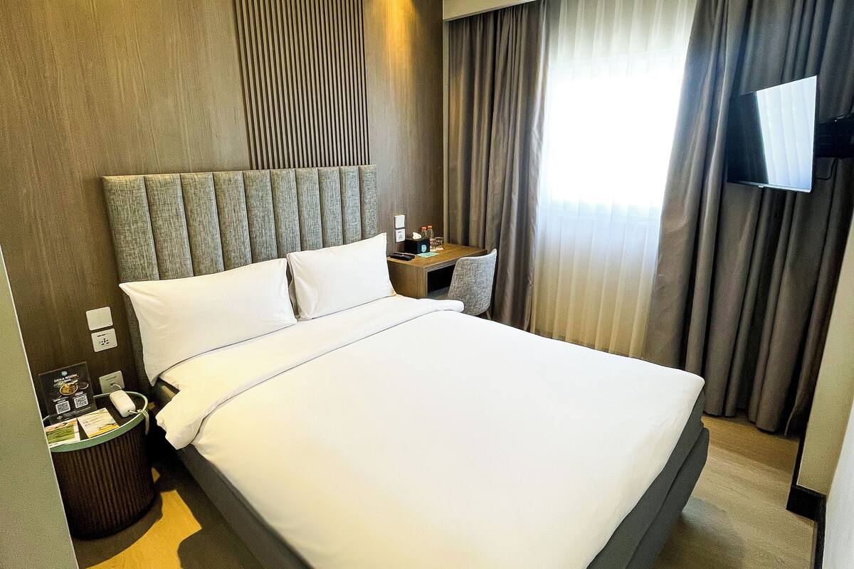 Quarto de hotel com uma cama grande, mesa de cabeceira com abajur, TV na parede e janela com cortinas.