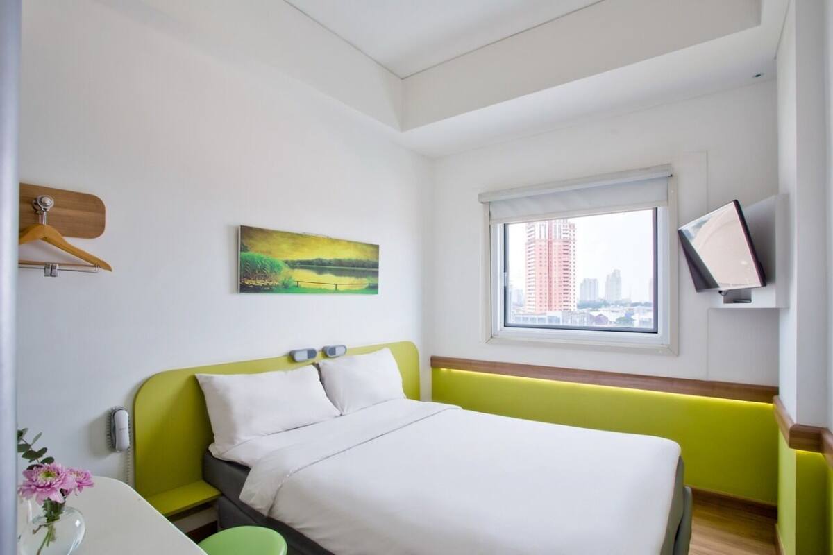 Quarto de hotel com cama, janela para a vista da cidade, quadro na parede e televisão montada.