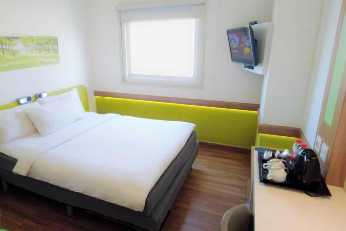 Quarto de hotel com cama, escrivaninha, cadeira, televisão e janela.