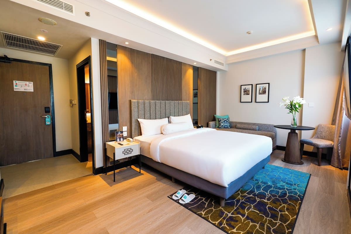 Quarto de hotel moderno com cama grande, mesa de cabeceira, um pequeno espaço de estar e vista para o corredor.