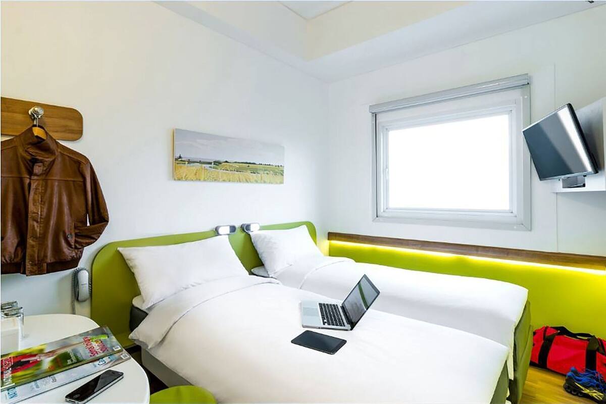 Quarto de hotel com cama, laptop, televisão, janela e um quadro na parede.