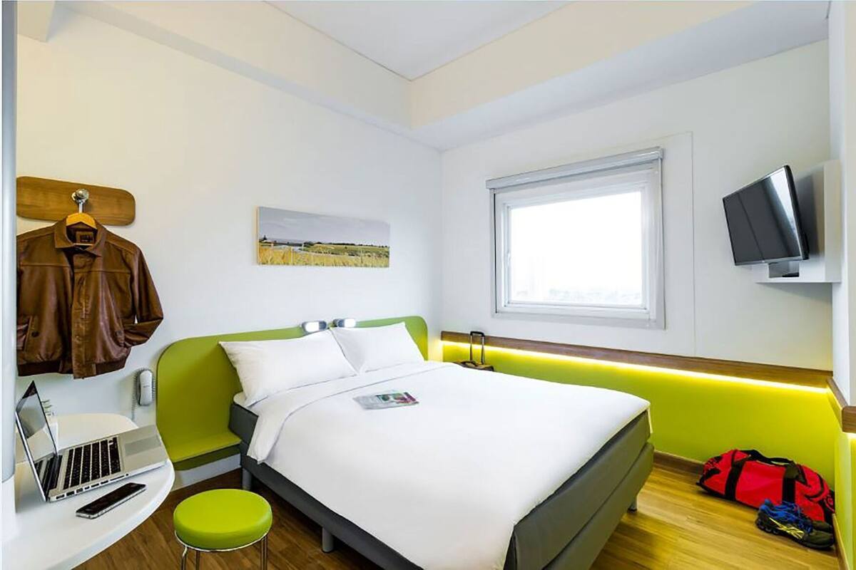 Quarto de hotel com cama, escrivaninha com um laptop, cadeira, televisão e janela.