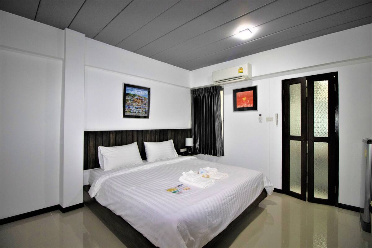 Quarto de hotel com cama grande, paredes brancas, cabeceira escura, ar condicionado, quadro emoldurado e porta com painéis de vidro jateado.