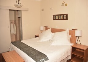 Uma cama bem arrumada com manta cinza, cabeceira de madeira, mesas de cabeceira com abajures e um quadro na parede.