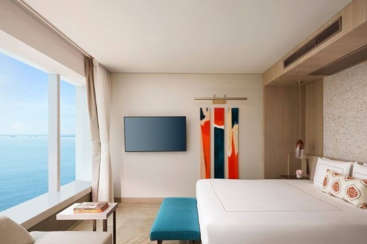 Quarto de hotel com uma cama grande, uma mesinha de cabeceira, um banco, uma TV na parede e uma janela com vista para o mar.