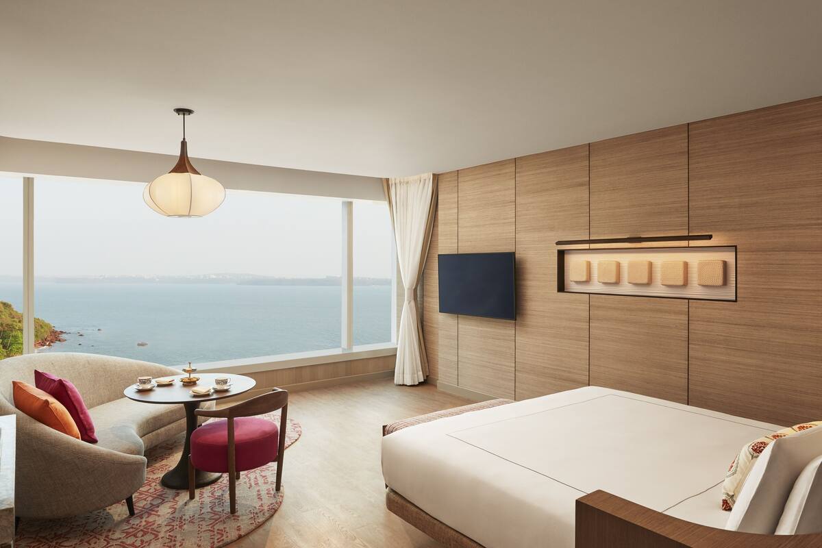 Quarto de hotel moderno com uma janela grande que oferece vista para o mar, uma cama, um espaço de estar com uma mesa e uma TV de tela plana fixada na parede.