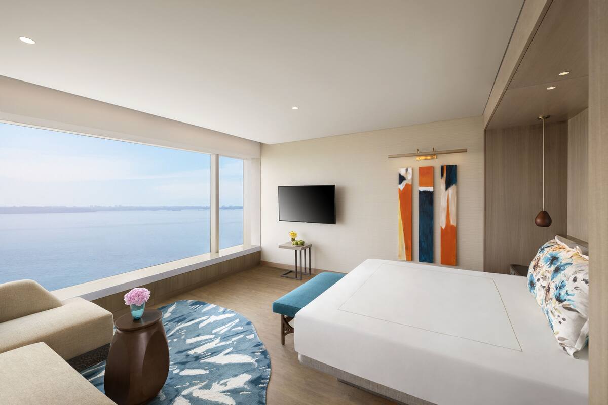 Quarto de hotel moderno com uma cama grande, um sofá, uma pequena mesa com um vaso, uma televisão de tela plana e vista para o mar.