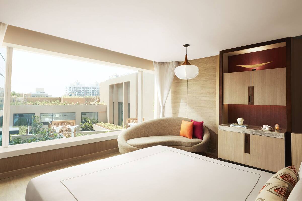 Quarto de hotel moderno com uma cama grande, área de estar com uma poltrona, vista para a cidade e uma janela ampla.
