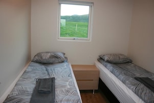 Quarto pequeno com duas camas individuais, cada uma com roupa de cama e almofadas estampadas. Uma pequena mesa de cabeceira de madeira fica entre as camas. Uma janela sobre as camas oferece vista para uma paisagem verde.