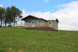 Uma cabana térrea com deck de madeira e um pequeno alpendre, cercada por um campo gramado e árvores.