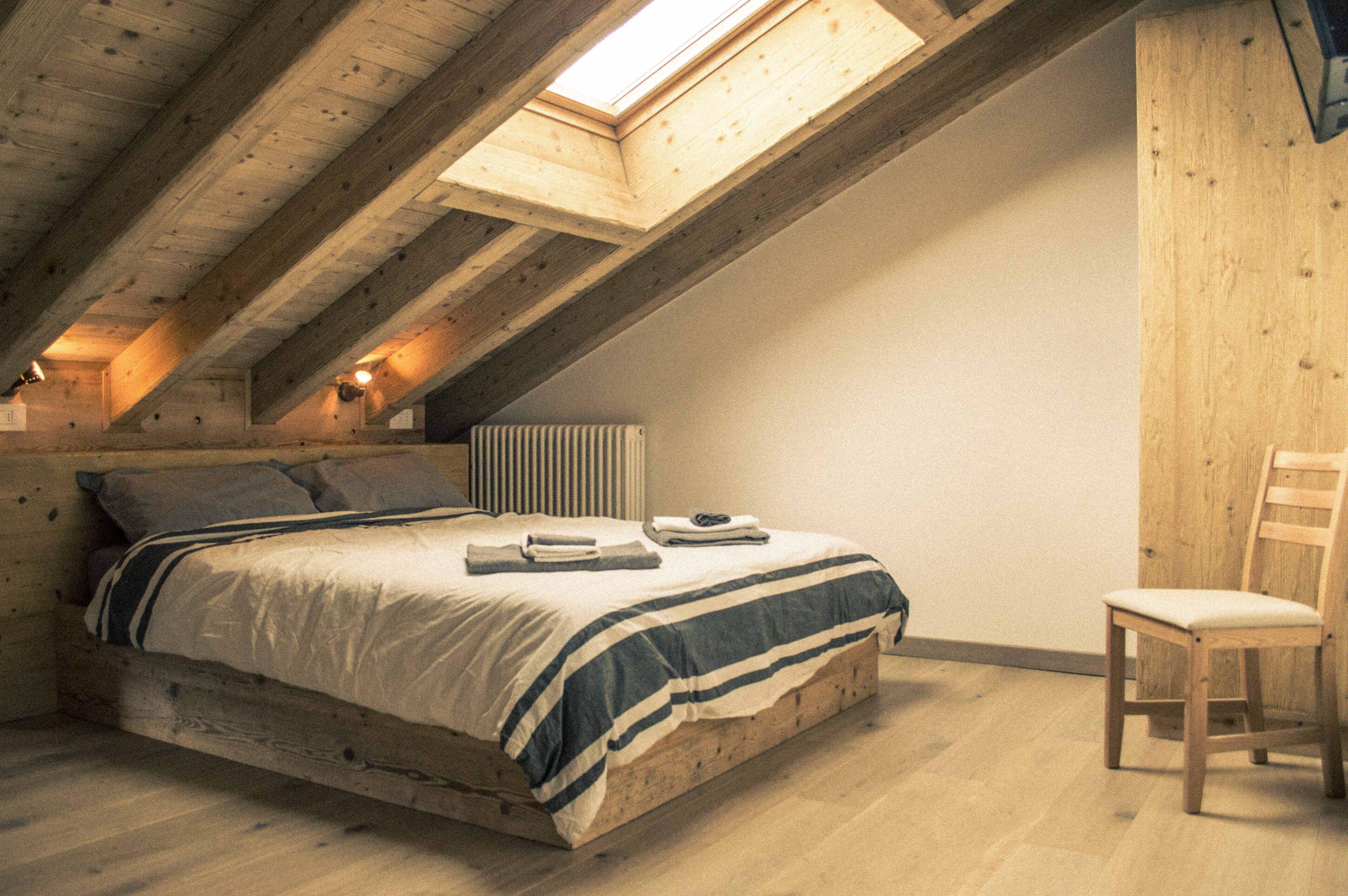 Quarto em estilo loft com teto inclinado, vigas de madeira, cama com roupa de cama listrada, radiador e uma cadeira.