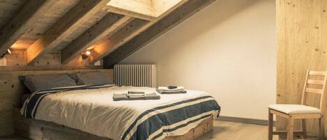 Quarto em estilo loft com teto inclinado, vigas de madeira, cama com roupa de cama listrada, radiador e uma cadeira.