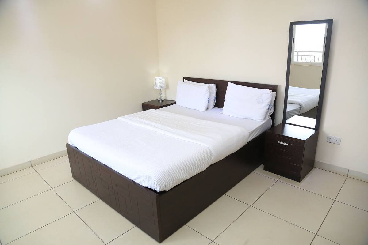 Cama bem arrumada com lençóis brancos em quarto com um espelho grande e mesa de cabeceira com abajur.