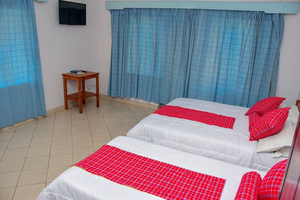Quarto com duas camas, uma mesinha de madeira e uma TV fixada na parede.