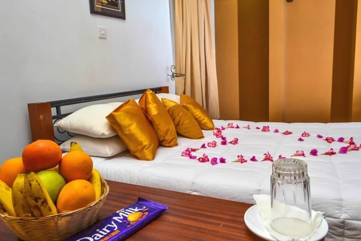Cama arrumada com almofadas douradas, uma cesta de frutas, uma barra de chocolate e um copo de leite sobre uma mesa de madeira.