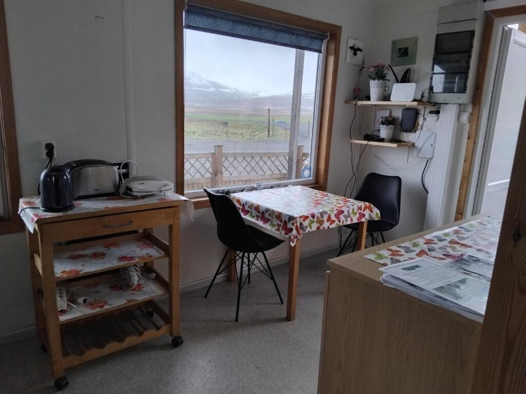 Quarto com escrivaninha de madeira, uma cadeira, janela com vista para a paisagem e uma estante com objetos.