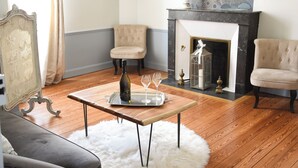 Uma sala de estar com piso de madeira, um tapete branco e fofo, uma mesa com uma bandeja contendo uma garrafa e copos, e duas cadeiras.