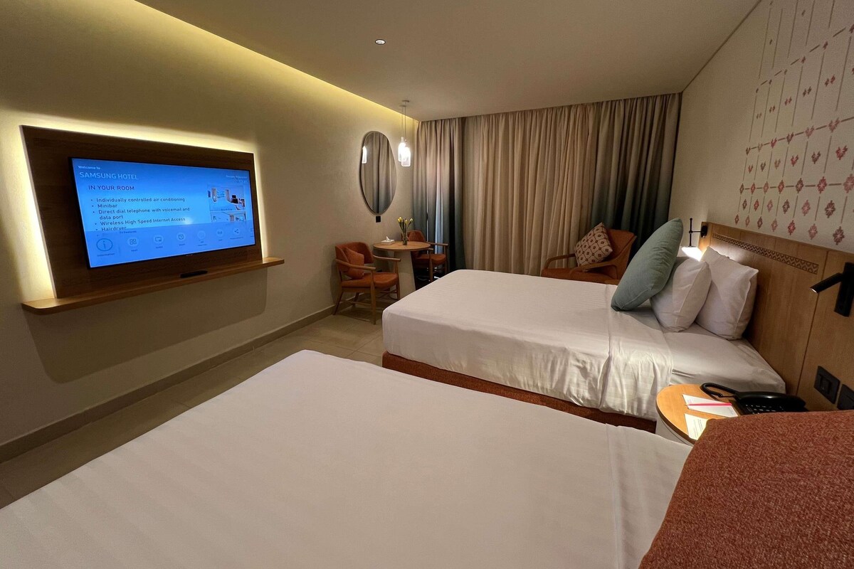 Quarto de hotel com duas camas, televisão fixada na parede, uma mesinha com uma cadeira e janela coberta por cortinas.