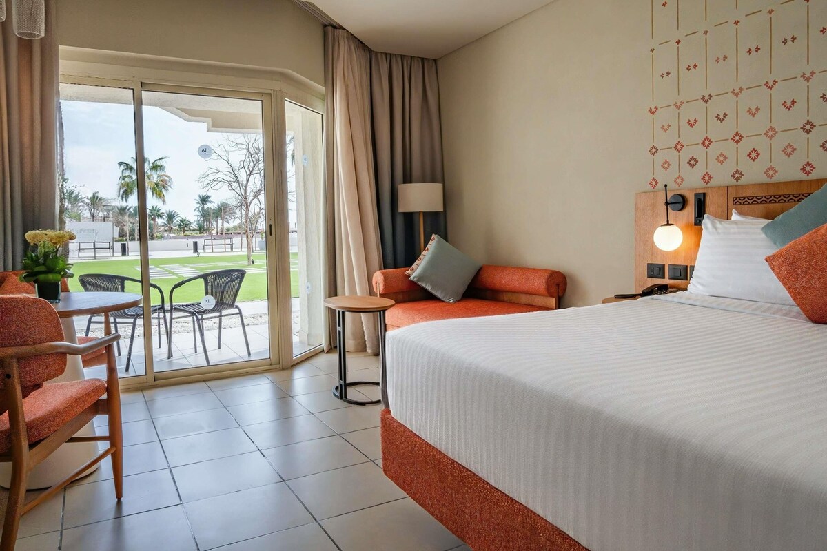 Quarto de hotel com cama, escrivaninha e vista para uma área verde ao ar livre.