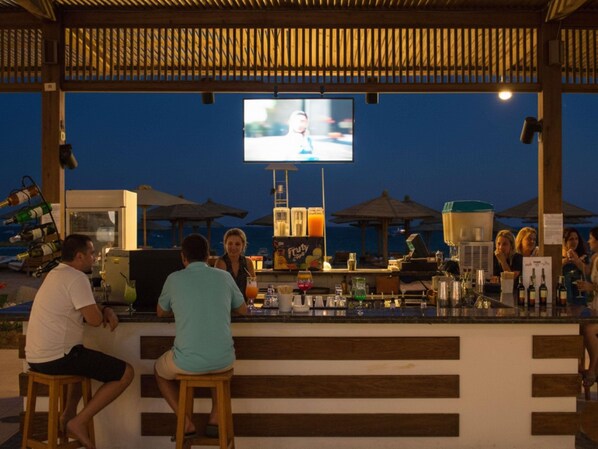 Uma área de bar com televisão, balcão, banquetes e vista para o mar.