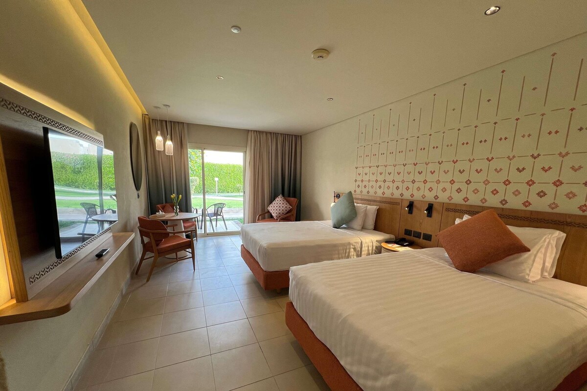 Quarto de hotel com duas camas, televisão, escrivaninha com cadeiras e vista para o exterior.