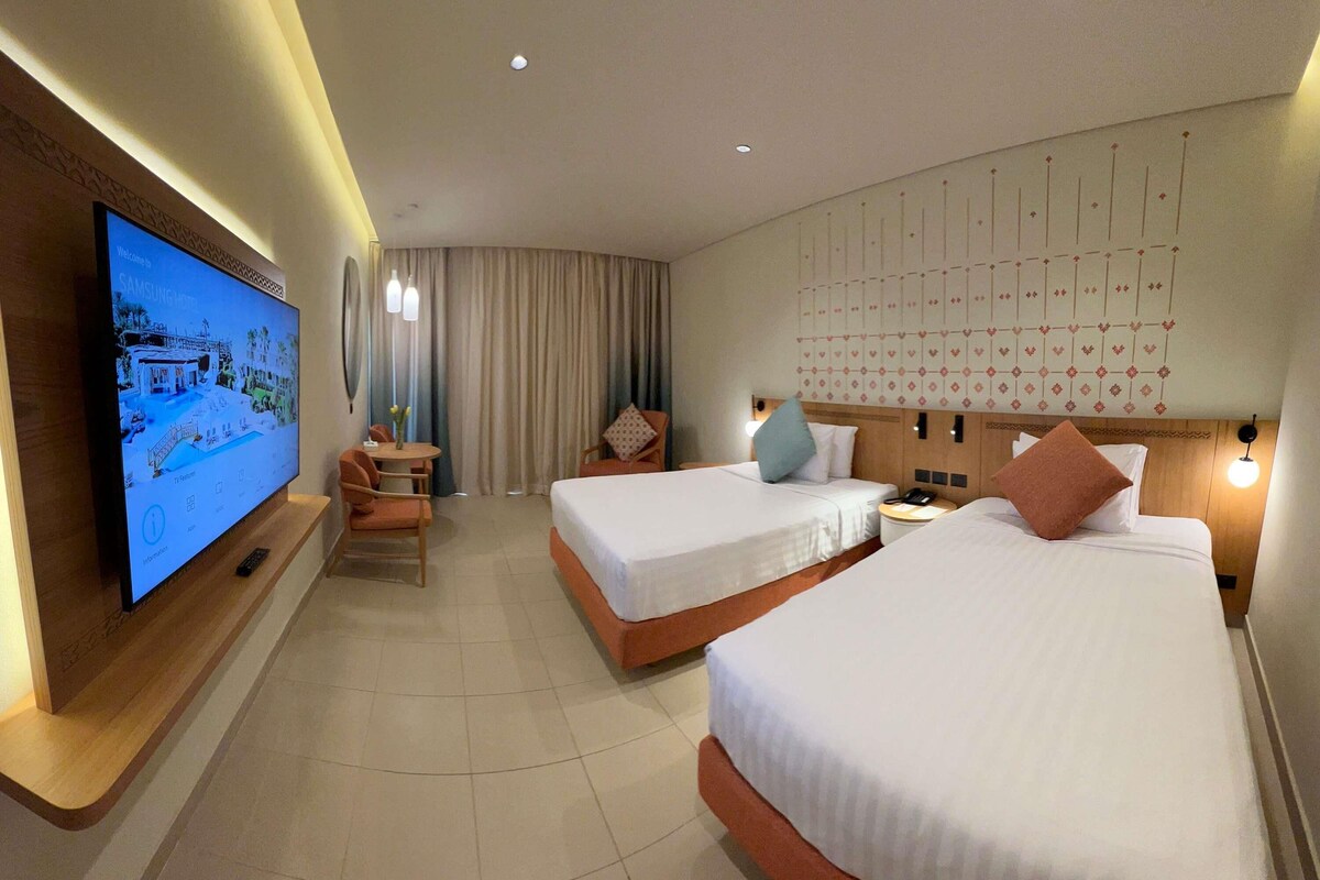 Quarto de hotel com duas camas, televisão na parede, secretária com cadeira e um painel decorativo.