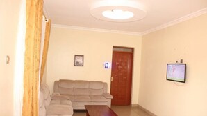 Uma sala de estar com um sofá bege, uma mesa de centro de madeira, uma televisão fixada na parede e uma porta ao fundo.