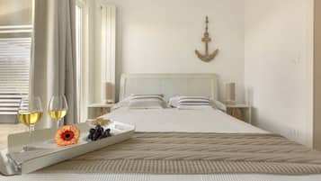 Uma cama bem arrumada com um cobertor bege, uma bandeja com uma flor e uvas, e duas taças de vinho branco sobre a mesa de cabeceira.