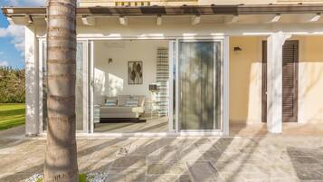 Uma casa moderna com varanda, uma grande porta de correr de vidro que dá para uma sala de estar com sofá e uma pintura, e uma palmeira em primeiro plano.