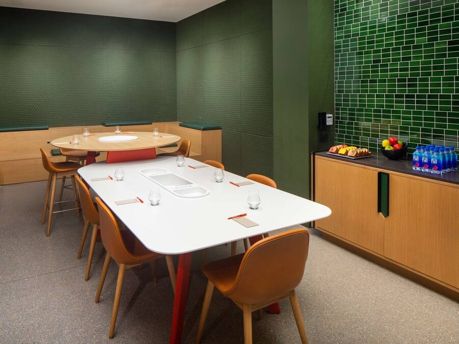 Sala de conferência com mesa redonda, cadeiras, quadro branco e uma pequena copa com fruteira e água engarrafada.