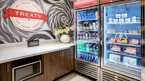Quarto de hotel com área de minibar, placa com a palavra 'Treats', um arranjo floral e duas geladeiras repletas de bebidas e lanches.