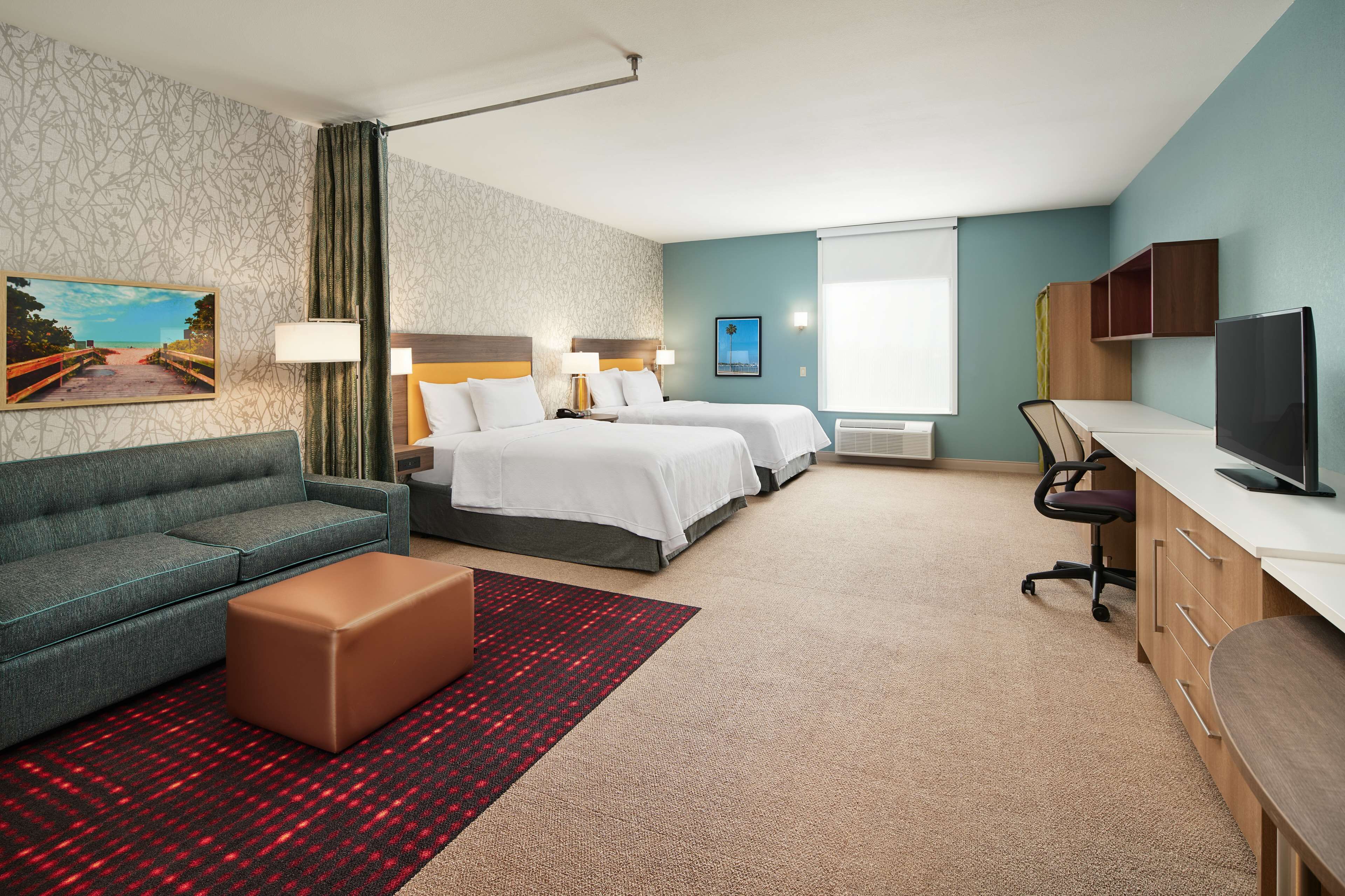 Quarto de hotel com duas camas, um sofá, uma escrivaninha, uma TV e um quadro na parede.