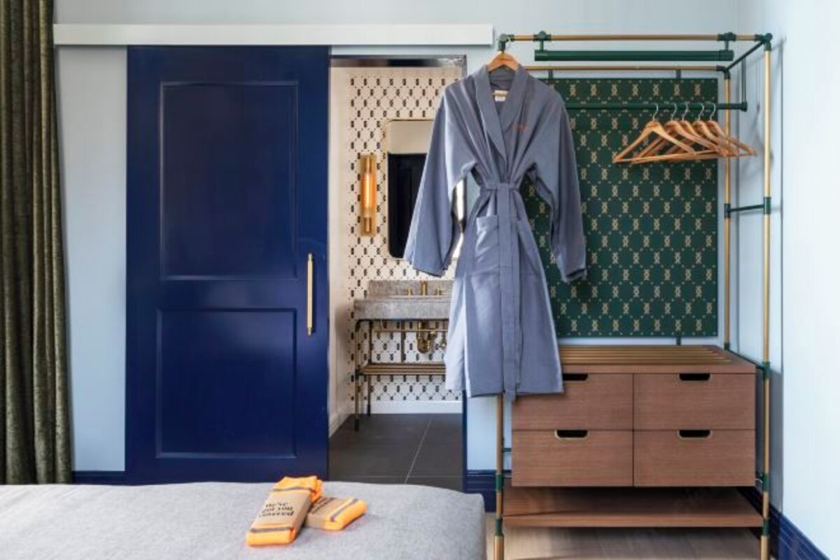 Quarto com cama, cómoda de madeira, roupão pendurado em um cabide e banheiro visível por uma porta aberta.