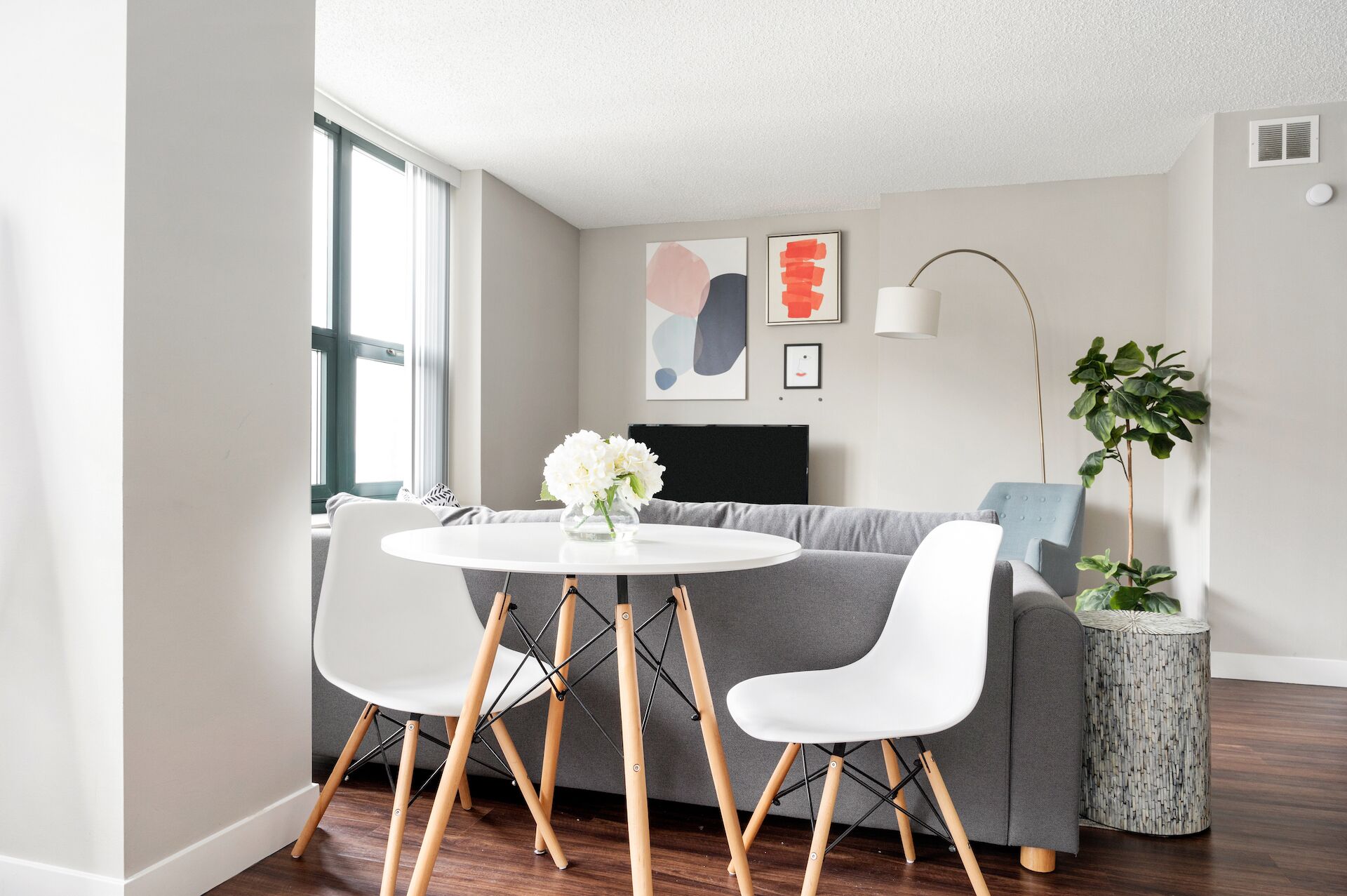Uma sala de estar moderna com uma mesa branca arredondada, duas cadeiras brancas, um sofá cinza e uma janela grande.