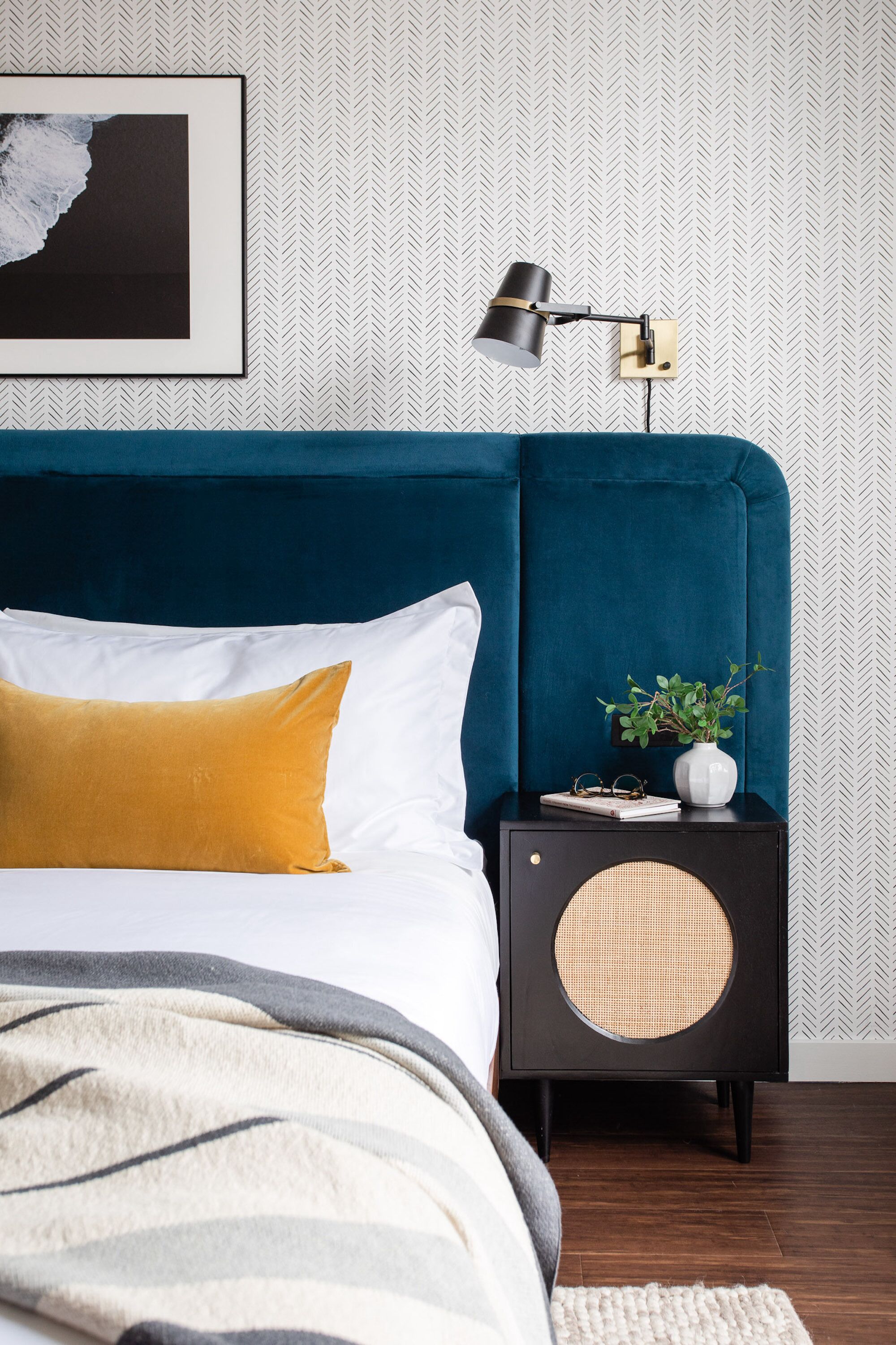Quarto com cabeceira estofada em tons de verde-água, cama branca e mostarda, mesa de cabeceira com uma planta e um quadro na parede.