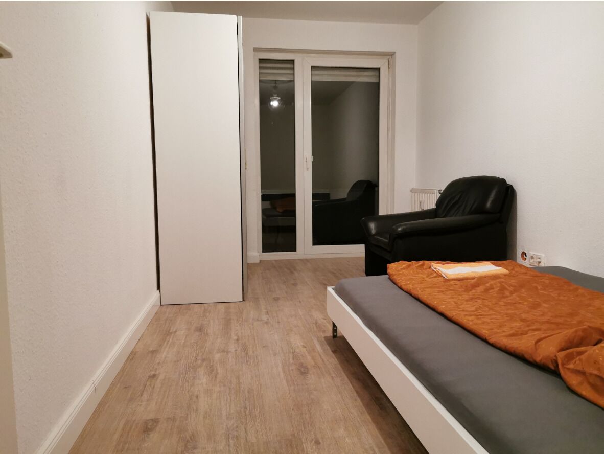 Quarto com cama, uma cadeira, guarda-roupa e porta deslizante para uma varanda.