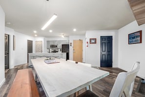 Uma cozinha moderna com eletrodomésticos de aço inoxidável, uma área de jantar com uma mesa comprida e cadeiras, e uma porta azul escura.