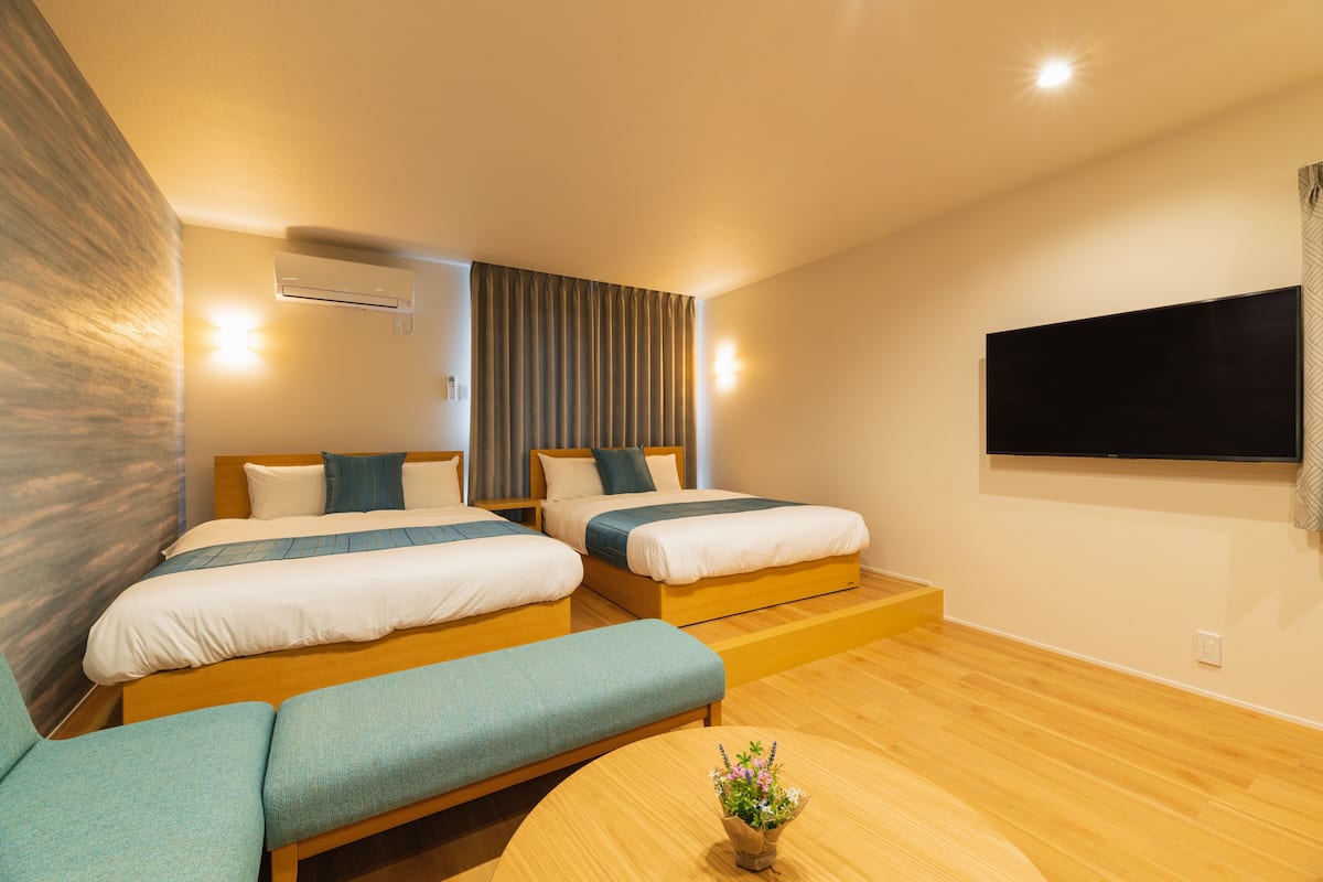 Quarto de hotel com duas camas, um sofá, uma TV de tela plana e uma mesinha com um vaso de flores.