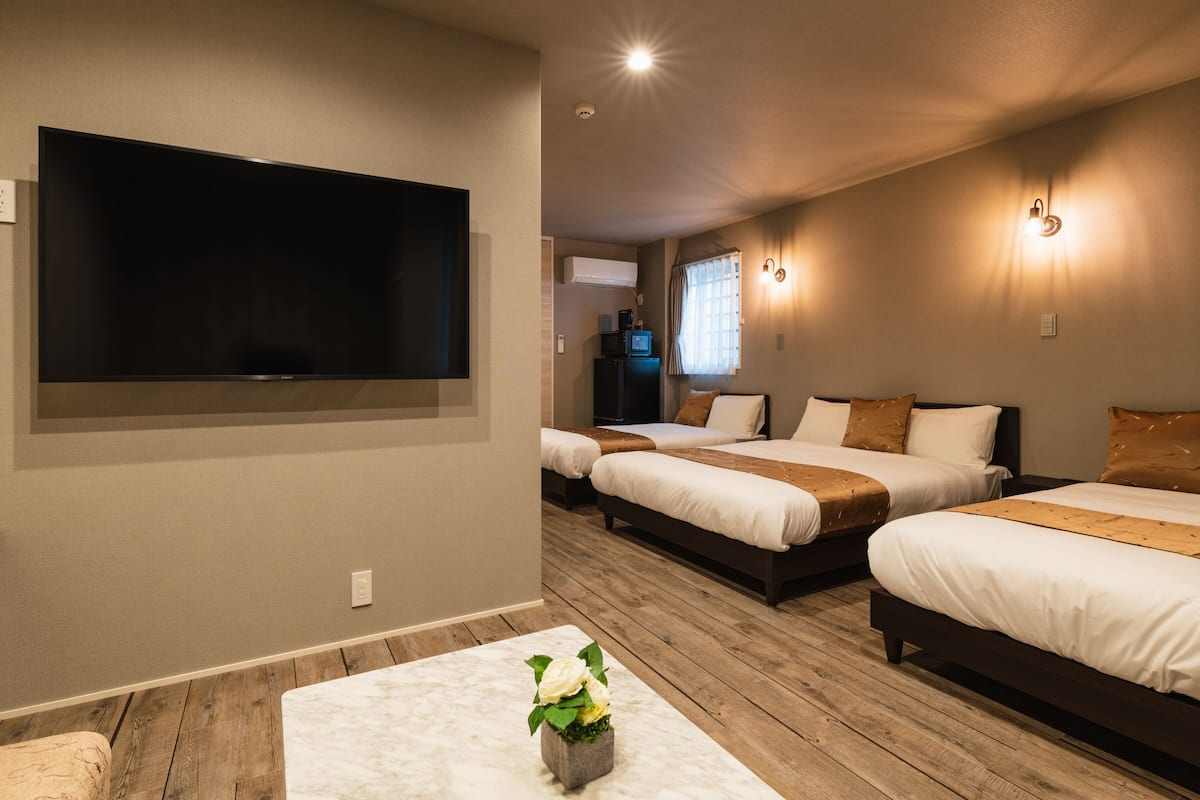 Quarto de hotel com três camas, televisão de tela plana, uma mesinha com um vaso e janela com cortinas.