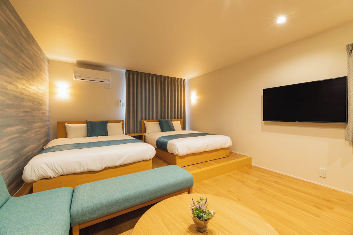 Quarto de hotel com duas camas, um sofá, uma TV de tela plana e uma mesinha com um vaso de flores.