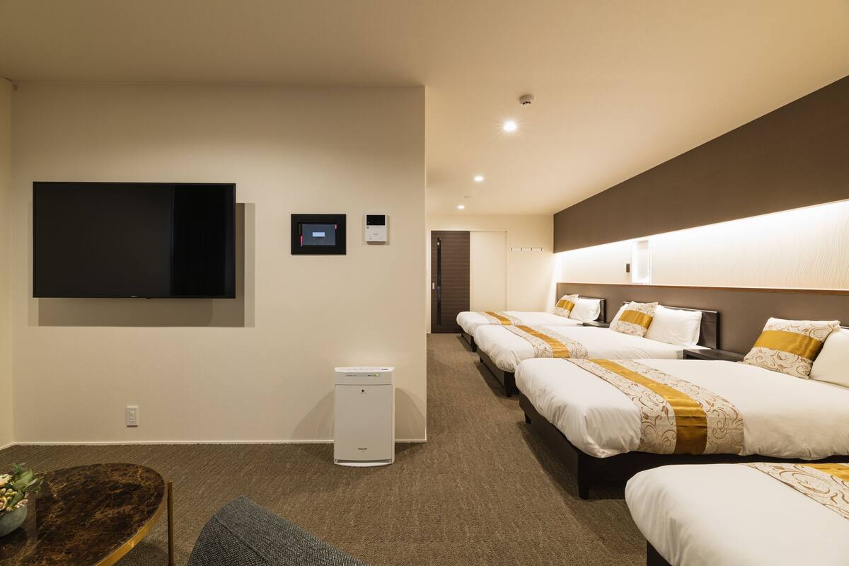 Quarto de hotel com uma TV de tela plana grande presa na parede, um purificador de ar compacto e uma cama com roupa de cama branca e detalhes dourados.