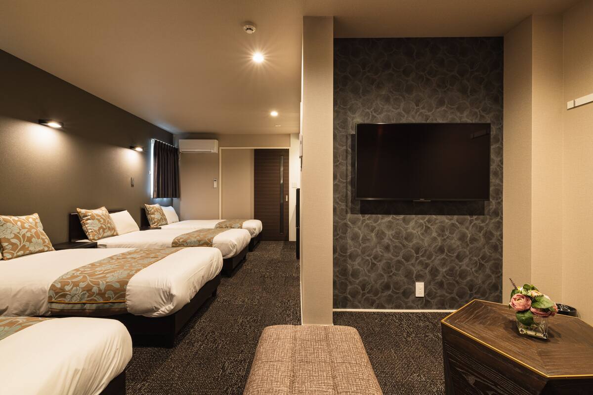 Quarto de hotel com quatro camas, uma televisão de tela plana na parede, uma pequena mesa de madeira com um vaso e piso coberto por carpete.