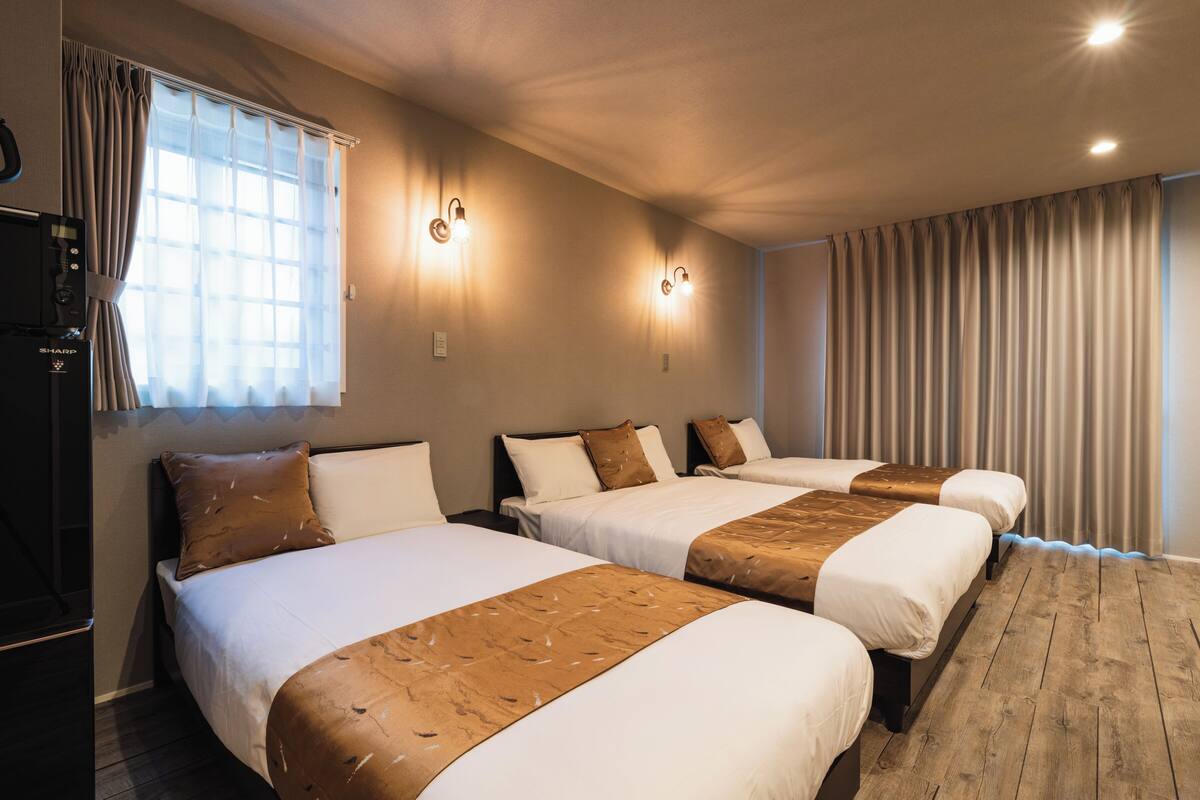 Quarto de hotel com três camas, piso de madeira e uma janela grande com cortinas.