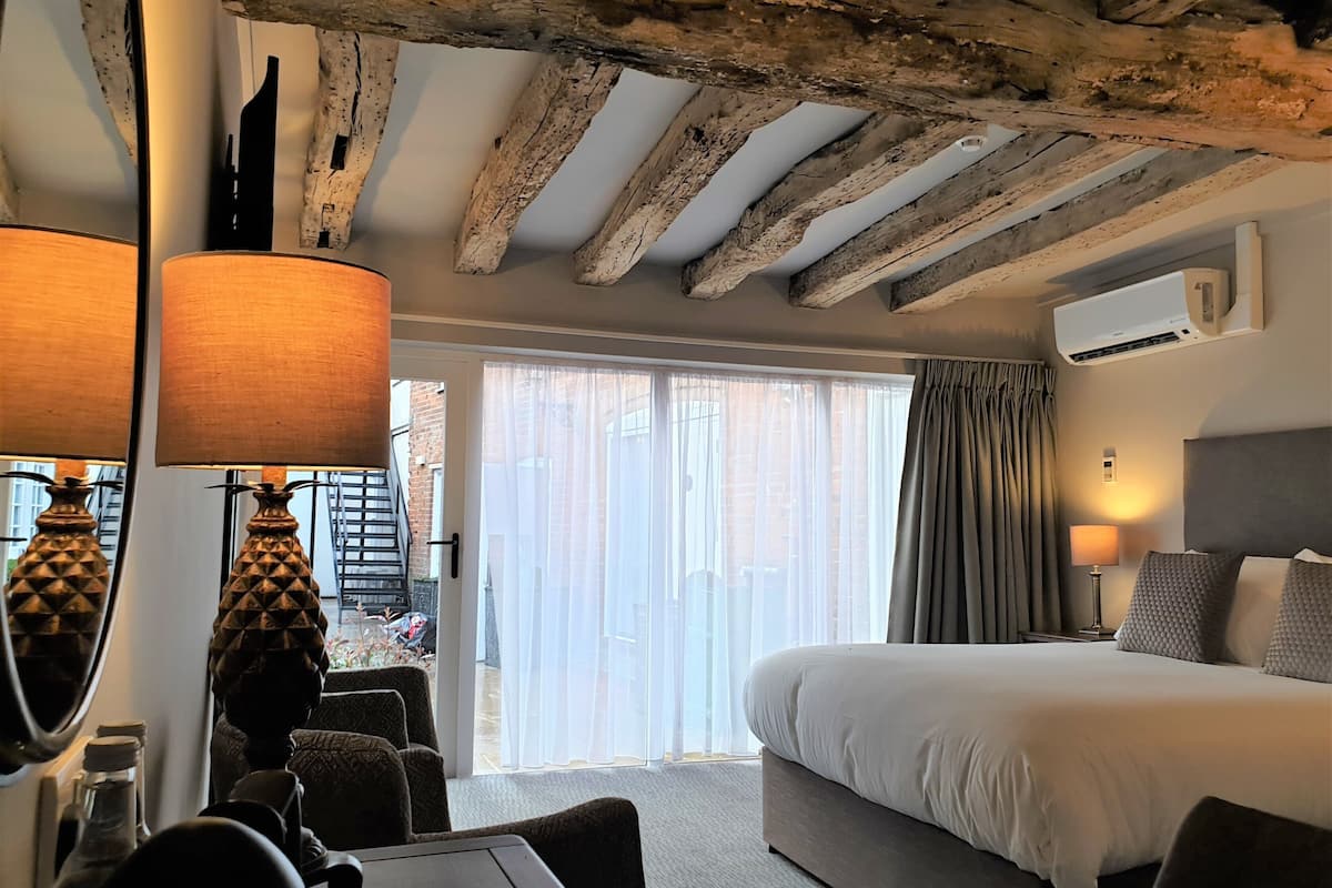 Quarto de hotel com cama, mesa, cadeiras, luminária, janela com cortinas e escada que dá para o exterior.