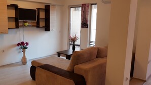 Uma sala de estar com sofá, uma mesinha, TV fixada na parede, um vaso com flores e uma janela com persianas.