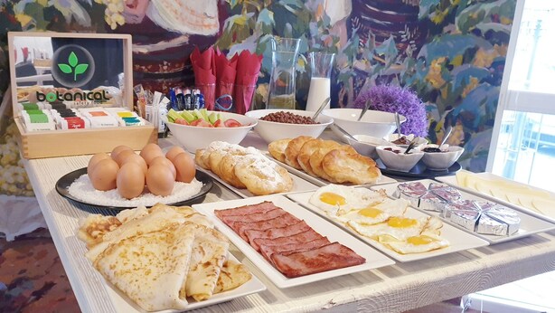 Buffet de café da manhã com diversos alimentos, incluindo ovos, presunto, pães e uma placa com a inscrição 'segurança alimentar botânica'.