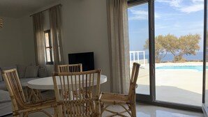 Um quarto com uma mesa redonda e quatro cadeiras, um sofá, uma televisão e uma janela grande com vista para a praia e piscina.