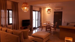 Uma sala de estar com sofá, banqueta, televisão e área de jantar com cadeiras e mesa. Há duas luminárias pendentes, uma porta de vidro deslizante e um ar condicionado na parede.