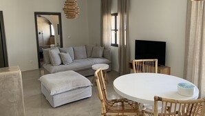 Uma sala de estar moderna com um sofá cinzento, um banquinho branco, cadeiras de vime, uma mesa redonda e uma televisão de tela plana.